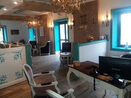 Quarto com piso de madeira, uma escrivaninha com um computador, uma cadeira e uma mesa com um cesto. Há uma parede de tijolos com uma janela e um aparelho de luz fixado na parede.
