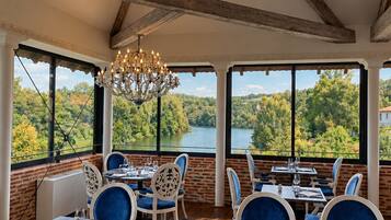 Uma sala de jantar com mesas e cadeiras, grandes janelas com vista para um lago e árvores, e um lustre.
