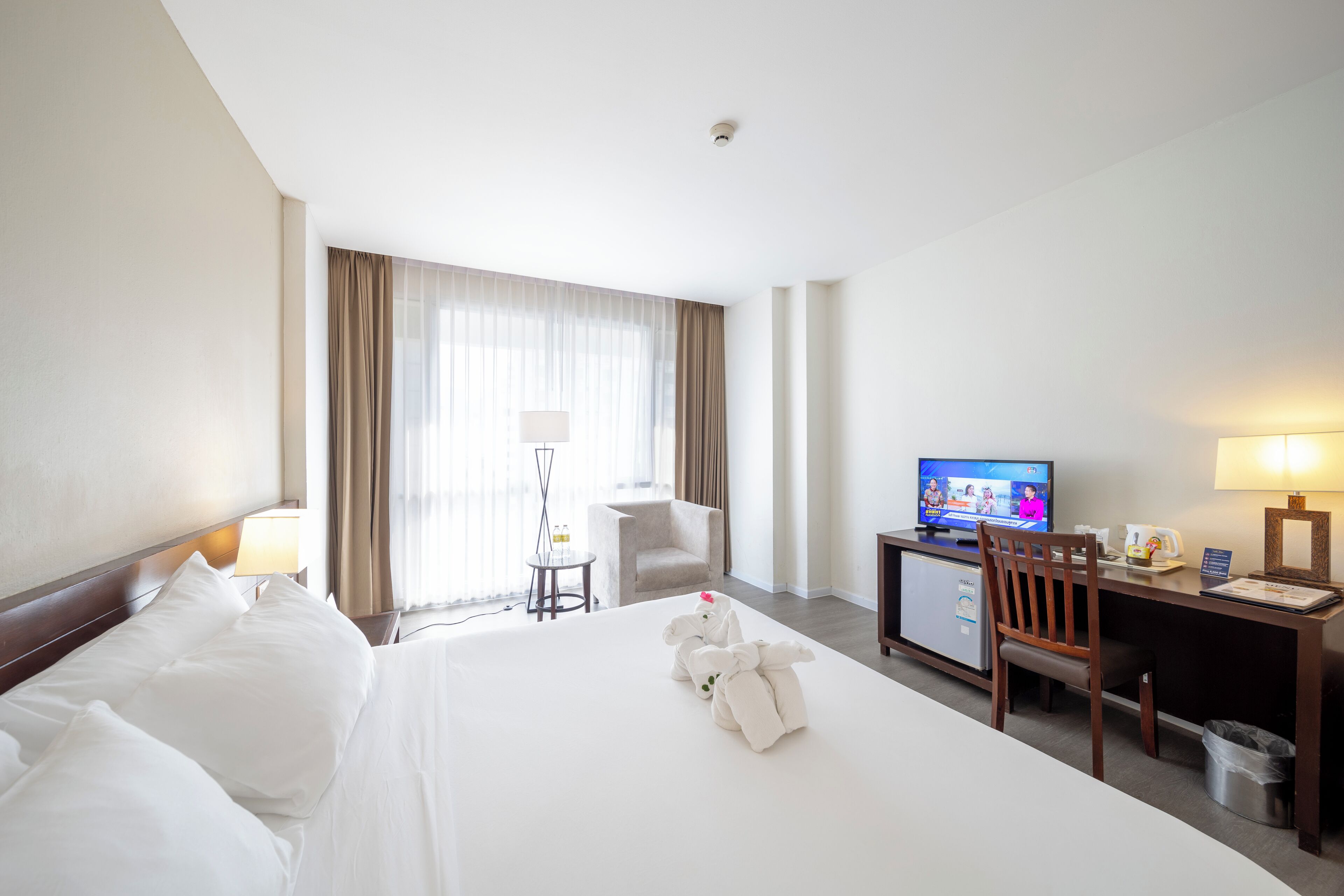 Quarto de hotel com uma cama grande, uma escrivaninha com televisão, uma cadeira e uma pequena mesa com abajur.