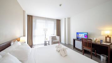 Quarto de hotel com uma cama grande, uma escrivaninha com televisão, uma cadeira e uma pequena mesa com abajur.