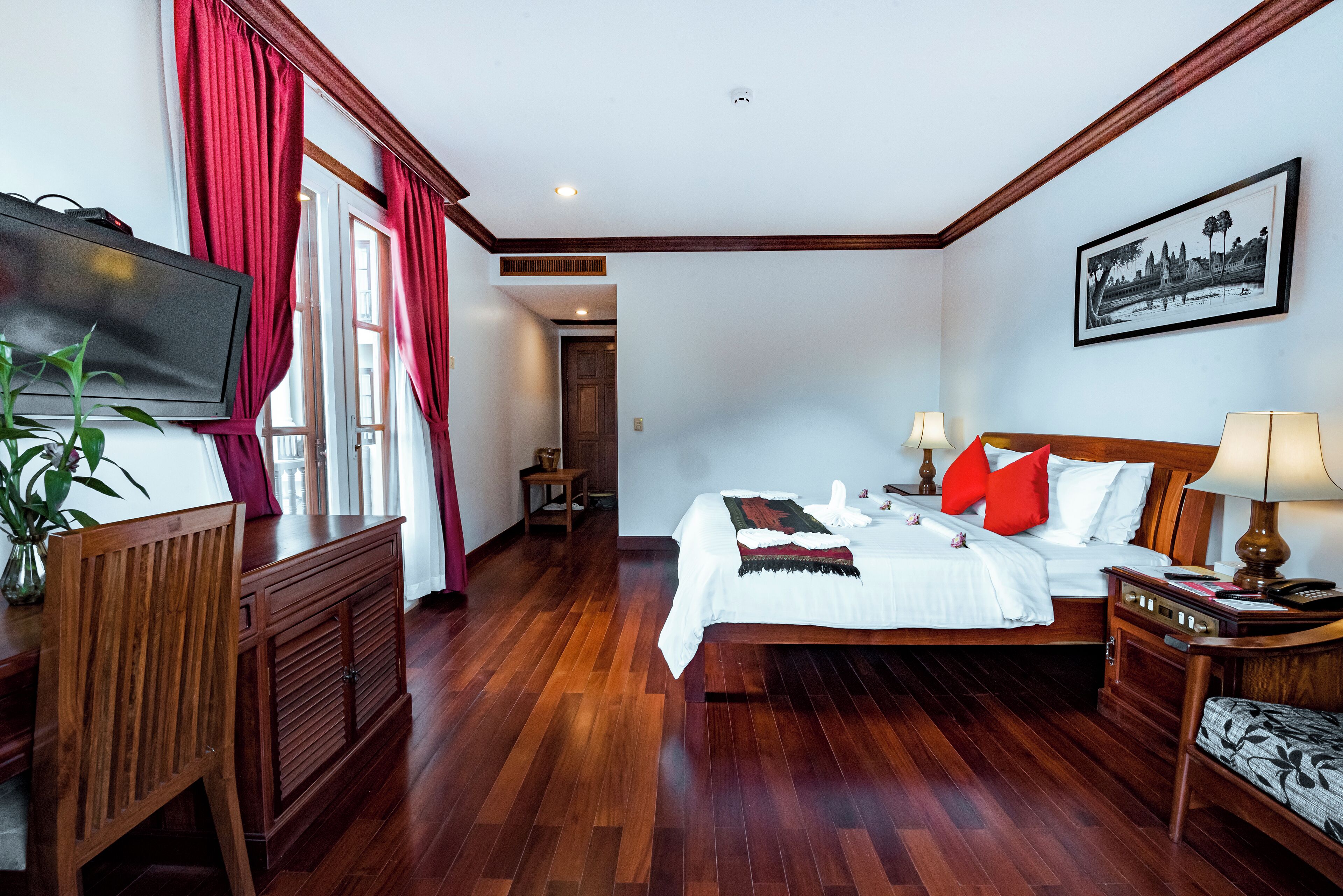 Quarto de hotel com cama de madeira, escrivaninha com televisão, mesa de cabeceira com abajur e quadro pendurado na parede.