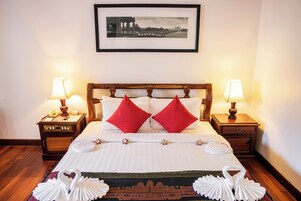 Uma cama bem arrumada com almofadas vermelhas, duas mesas de cabeceira com abajures, e um quadro na parede.