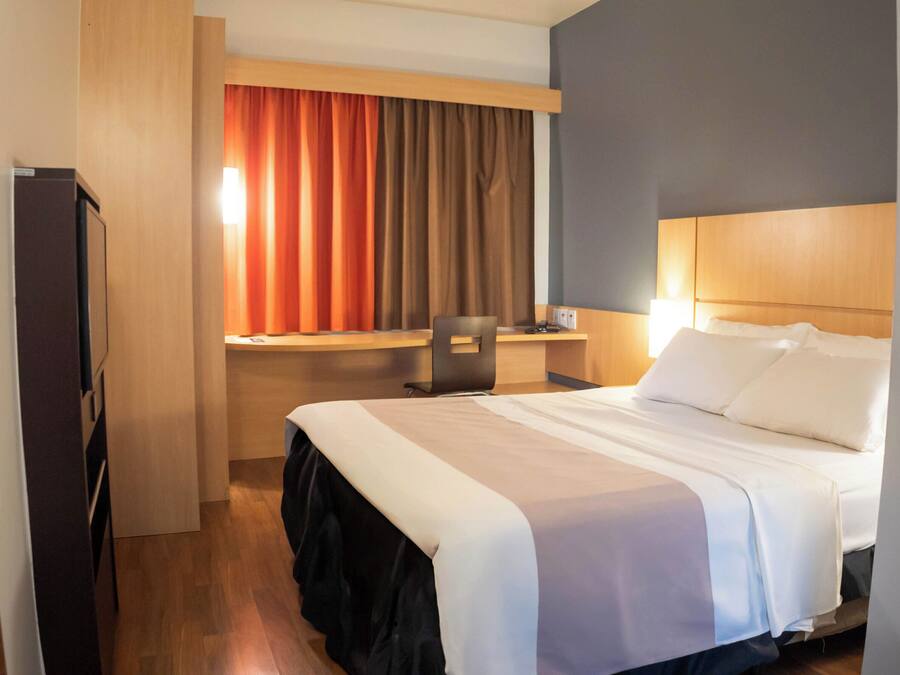 Quarto de hotel com cama, escrivaninha e um laptop, além de televisão.