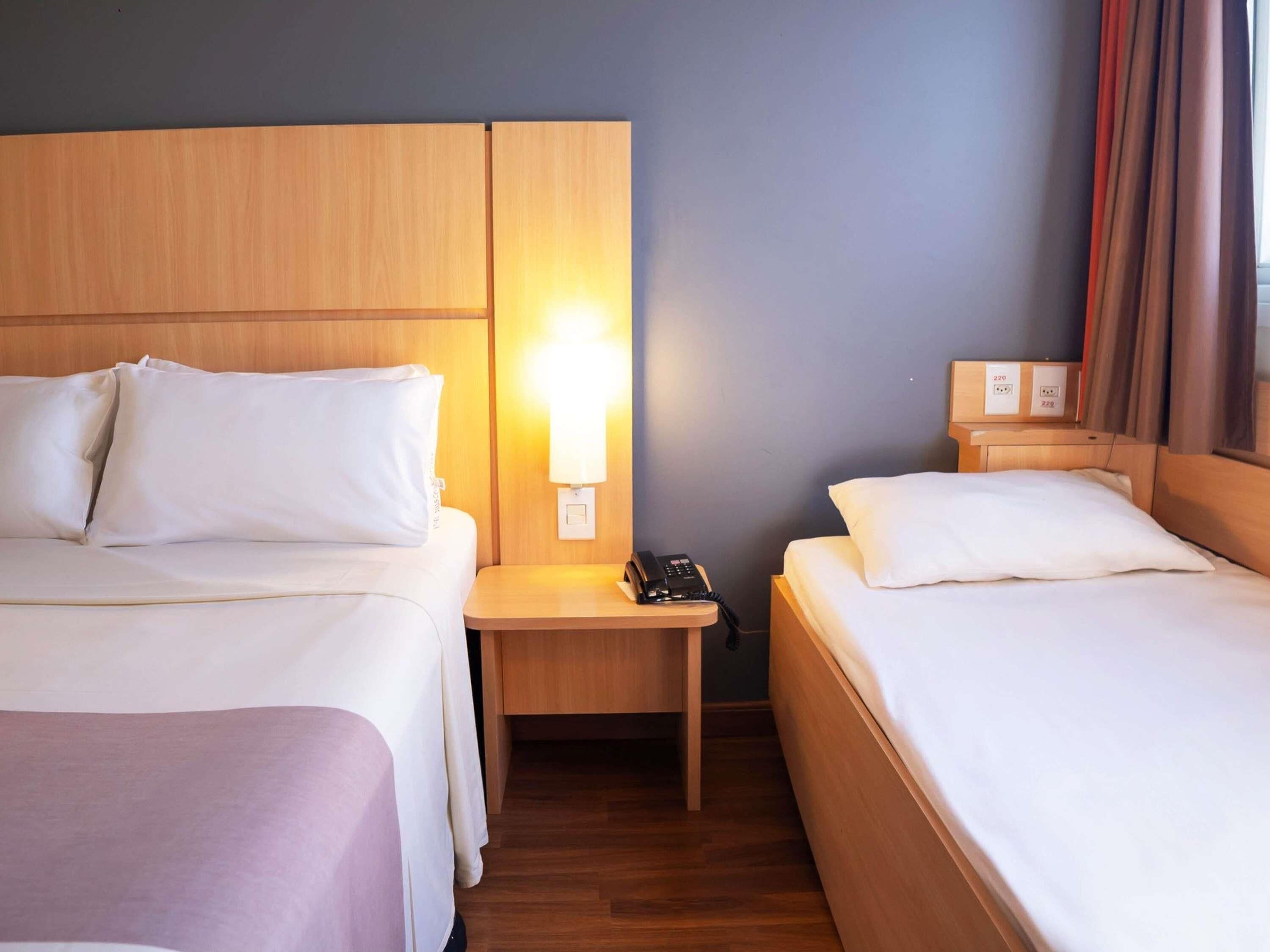 Quarto de hotel com duas camas, uma mesa de cabeceira com telefone e uma luminária na parede.