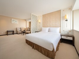 Quarto de hotel com uma cama grande, um espaço de estar com uma poltrona e uma mesa pequena, um painel decorativo na parede e um abajur.