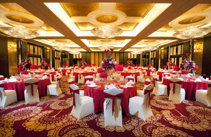 Salão de banquetes com mesas redondas, cadeiras brancas e toalhas de mesa vermelhas.