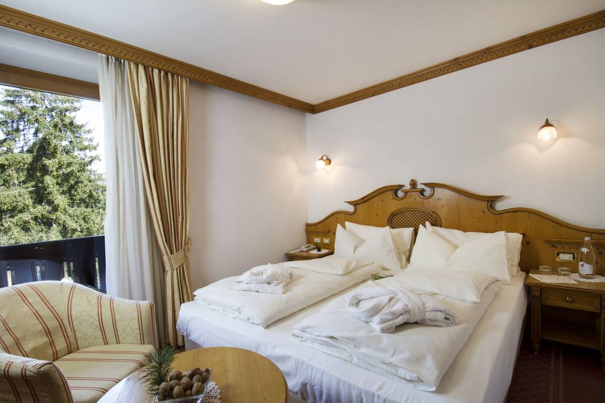 Quarto de hotel com cabeceira de madeira, duas camas com roupa de cama branca, mesa de cabeceira com abajur, uma cadeira e vista para árvores lá fora.