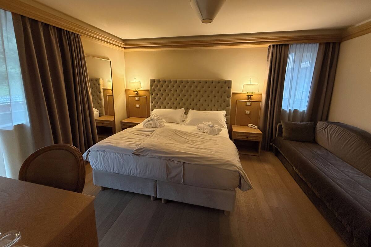 Quarto de hotel com uma cama grande, um sofá, uma escrivaninha e uma cadeira. Há duas mesas de cabeceira com luminárias e uma luminária no teto.