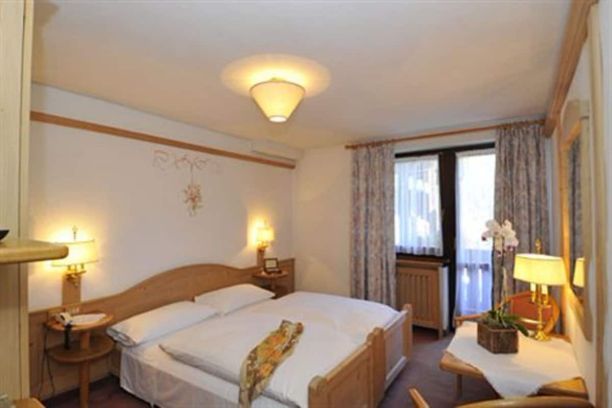 Quarto de hotel com uma cama grande, abajures, uma mesinha e uma janela com cortinas.