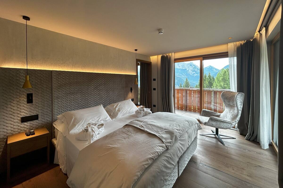 Quarto de hotel moderno com uma cama grande, mesa de cabeceira de madeira, uma cadeira e uma varanda com vista para as montanhas.