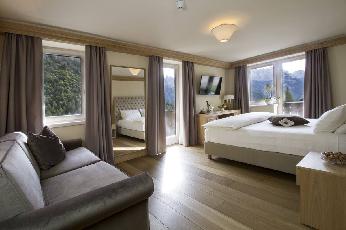 Quarto de hotel com cama, sofá, televisão e vista para as montanhas.