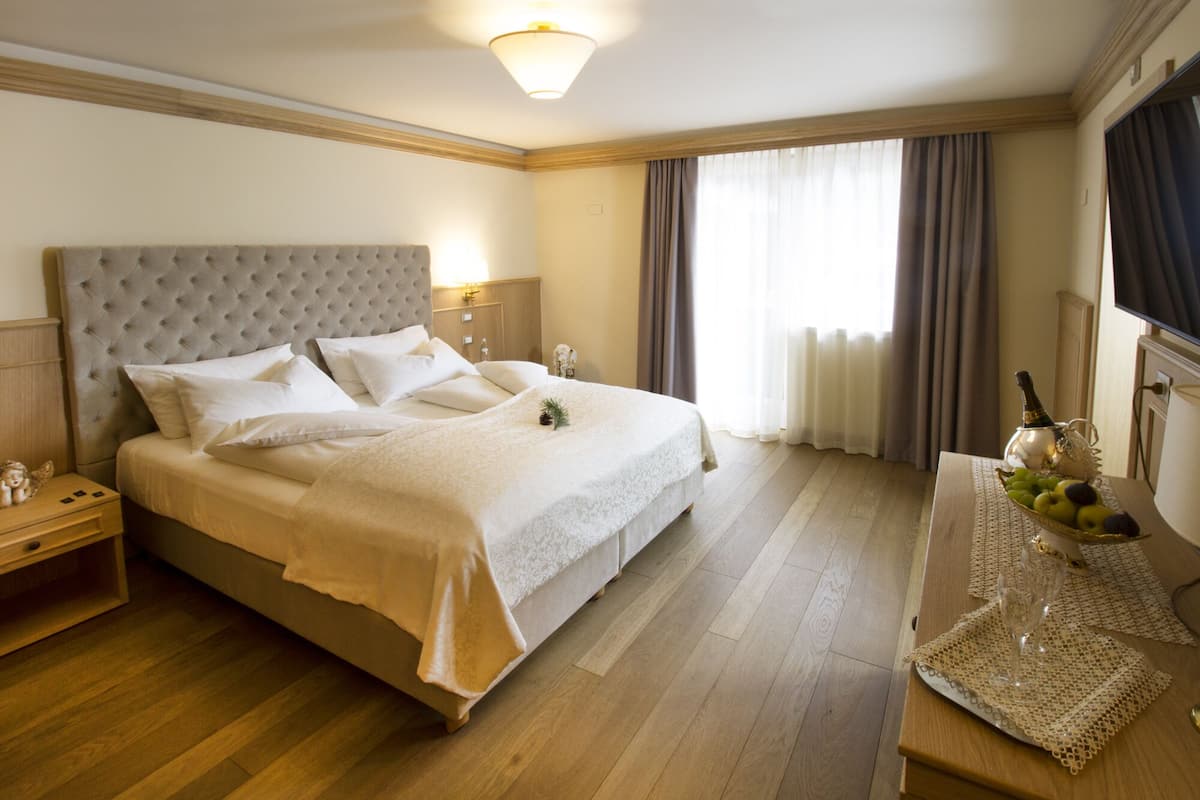 Quarto de hotel com uma cama grande, uma mesa de cabeceira de madeira, uma escrivaninha com uma tigela de frutas e uma garrafa, e uma televisão na parede.
