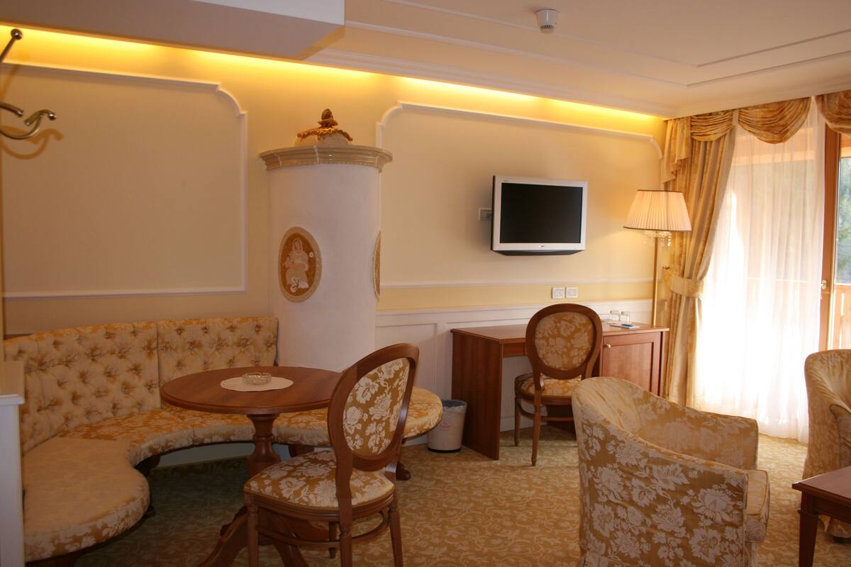 Quarto de hotel com sofá, mesa de jantar redonda, duas cadeiras, televisão, escrivaninha, luminária e janela com cortinas.