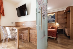 Quarto com parede de madeira, cama com colcha vermelha estampada, televisão fixada na parede e porta de vidro com adesivo de árvore.
