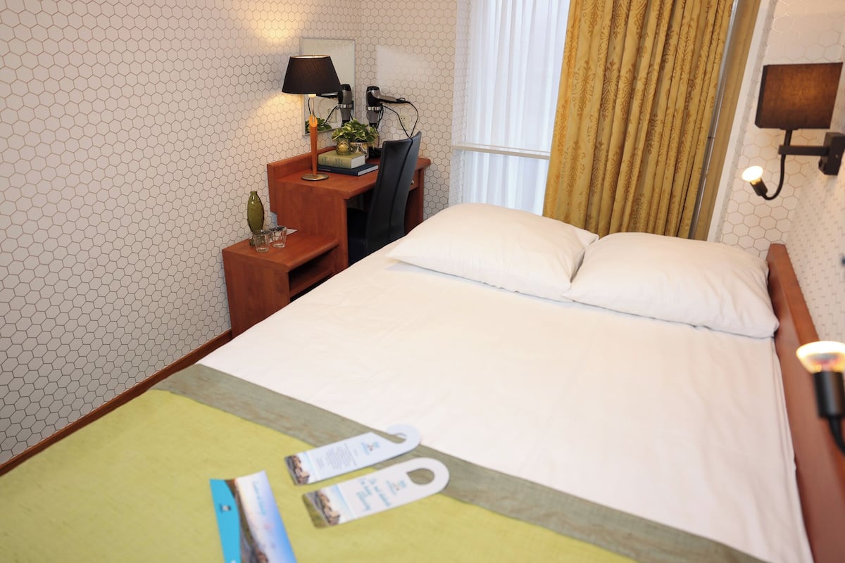 Quarto de hotel com uma cama bem arrumada, mesa de cabeceira com abajur, pequena escrivaninha com cadeira e janela com cortinas.