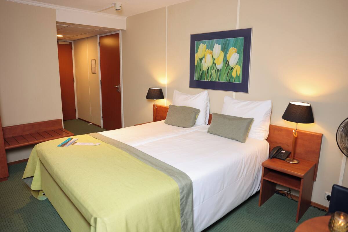 Quarto de hotel com cama, luminárias de cabeceira, mesa de cabeceira com telefone, um quadro na parede e porta para outro cômodo.
