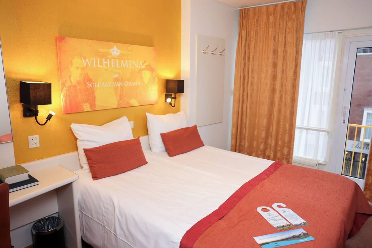 Quarto de hotel com cama, mesa de cabeceira, luminária, lixeira e janela com cortinas.
