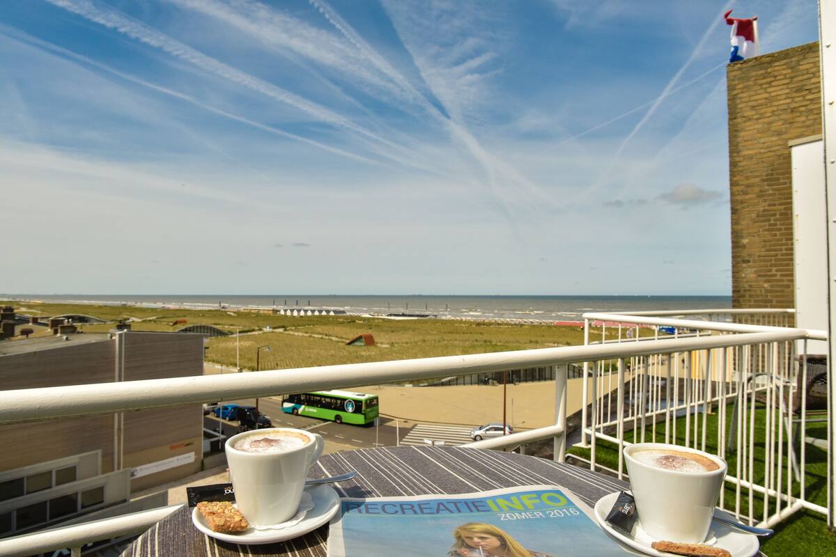 Varanda com vista para a praia, uma mesa com cardápio e duas chávenas de café.
