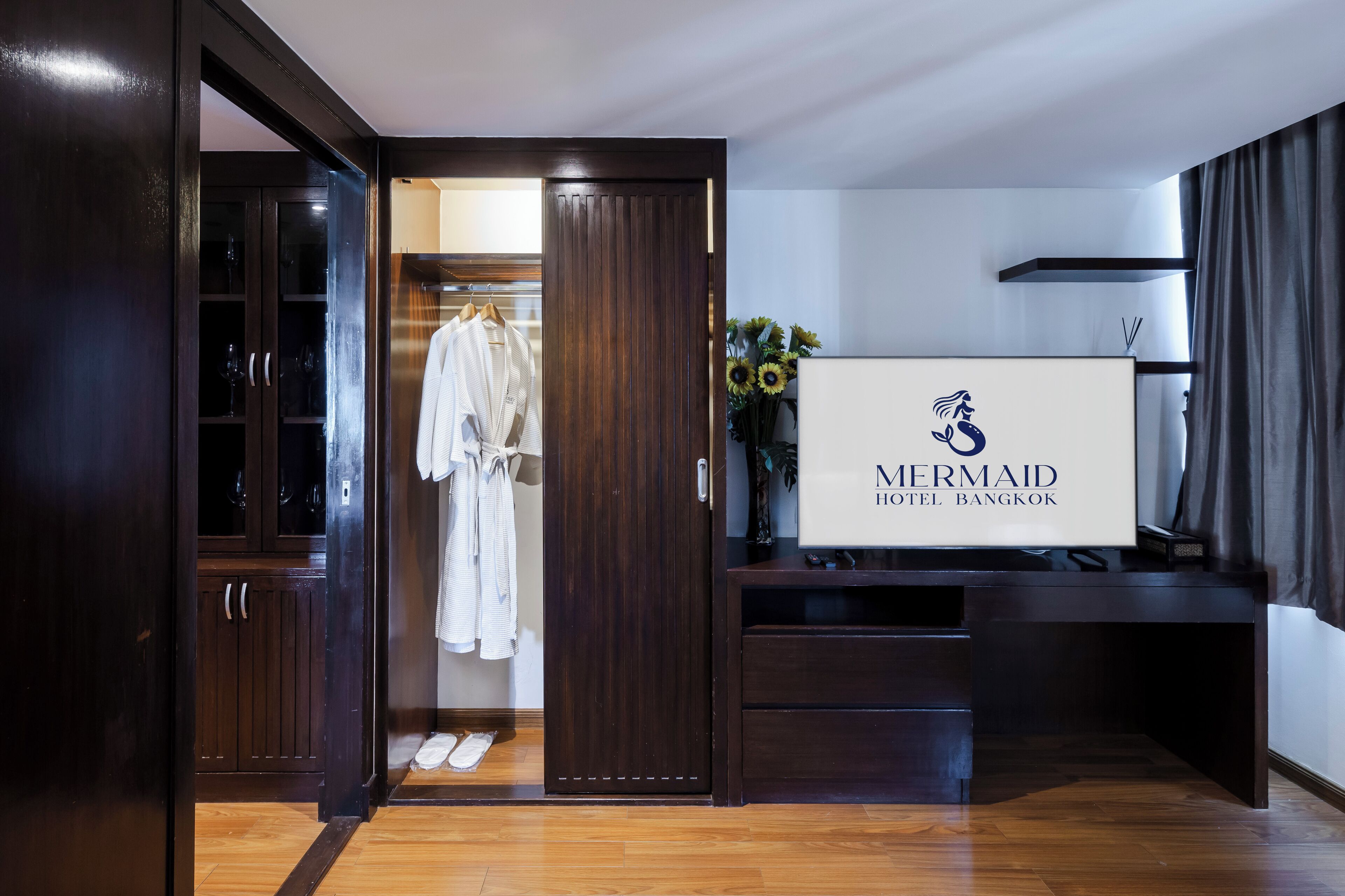 Quarto de hotel com um guarda-roupa de madeira, uma toucador com espelho e uma placa do Hotel Mermaid Bangkok.