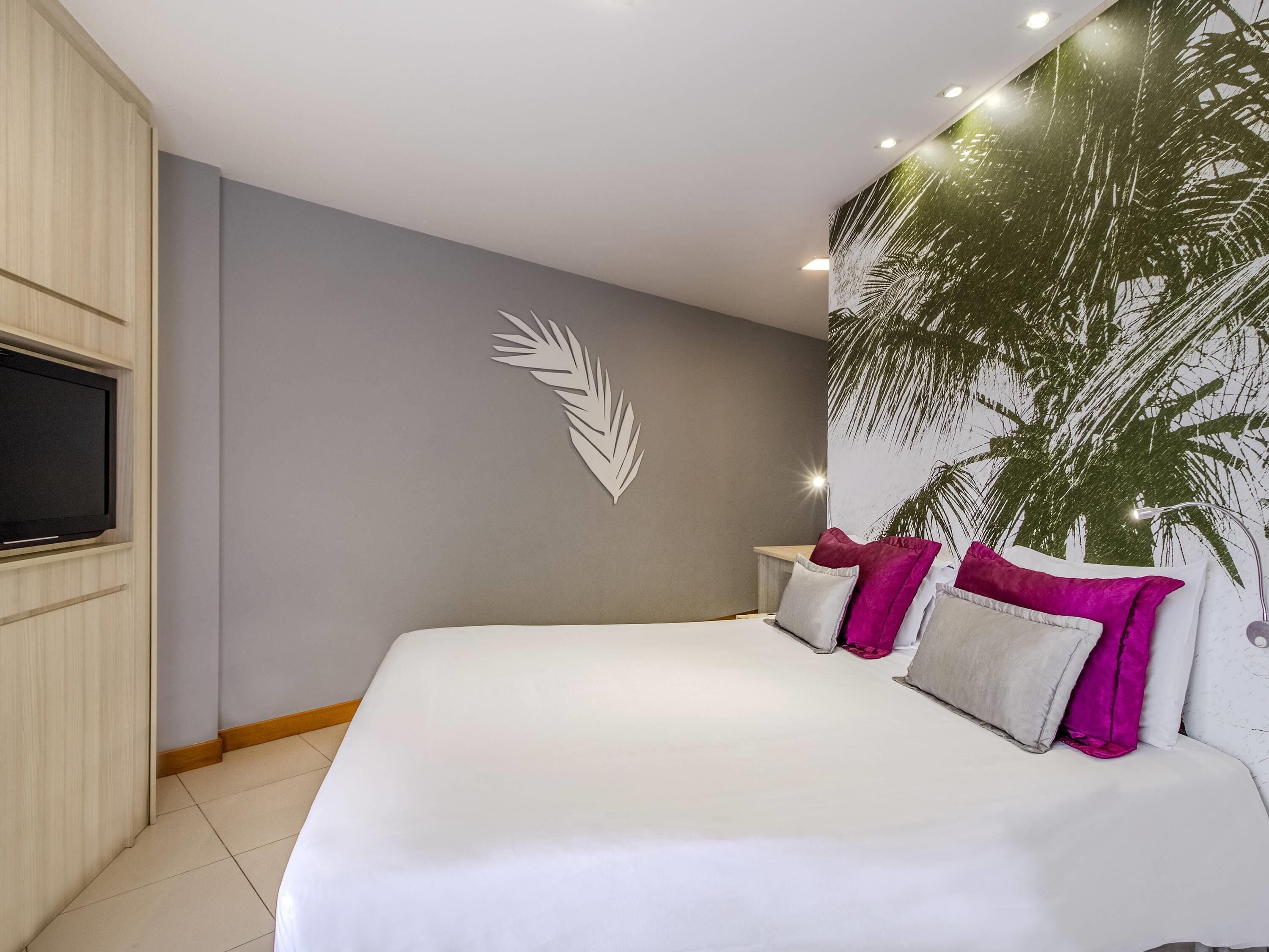Quarto com cama, TV, decoração em formato de folha na parede e um mural de palmeira.