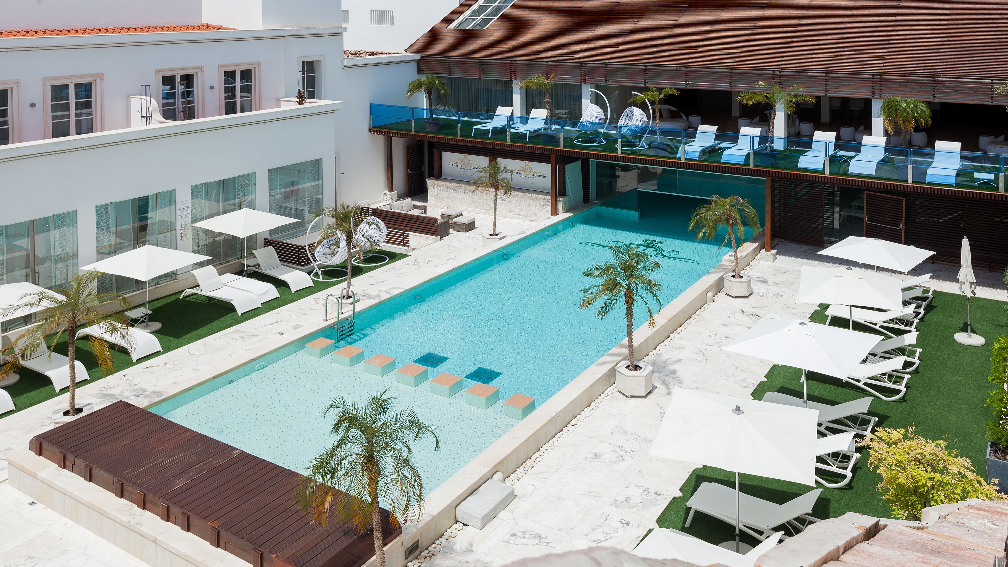 Piscina no terraço com espreguiçadeiras e guarda-sóis, cercada por um edifício branco com janelas amplas e uma varanda com mais espreguiçadeiras.
