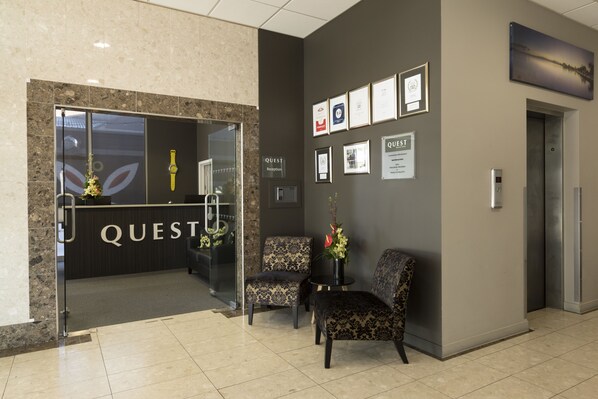 Uma área de recepção com uma placa 'QUEST', duas cadeiras estampadas, um vaso com flores e certificados emoldurados na parede.