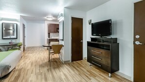 Quarto de hotel com piso de madeira, televisão em uma cômoda, uma pequena mesa de jantar com cadeiras, cama com travesseiro verde e banheiro com espelho e pia.