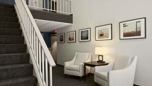 Um hall de hotel com uma escadaria, duas poltronas, uma mesa com abajur e quadros nas paredes.