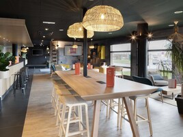 Um café moderno com uma mesa de madeira comprida, banquetas de bar e luminárias de vime penduradas.