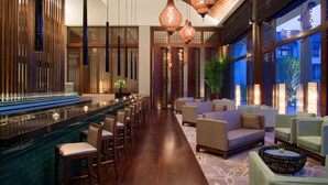 Uma área de bar moderna com um balcão comprido e vários bancos, um espaço de estar com sofás e cadeiras, e um teto com vigas de madeira e luminárias pendentes.
