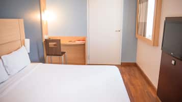 Quarto de hotel com cama, escrivaninha, cadeira, aparelho de ar condicionado e televisão.