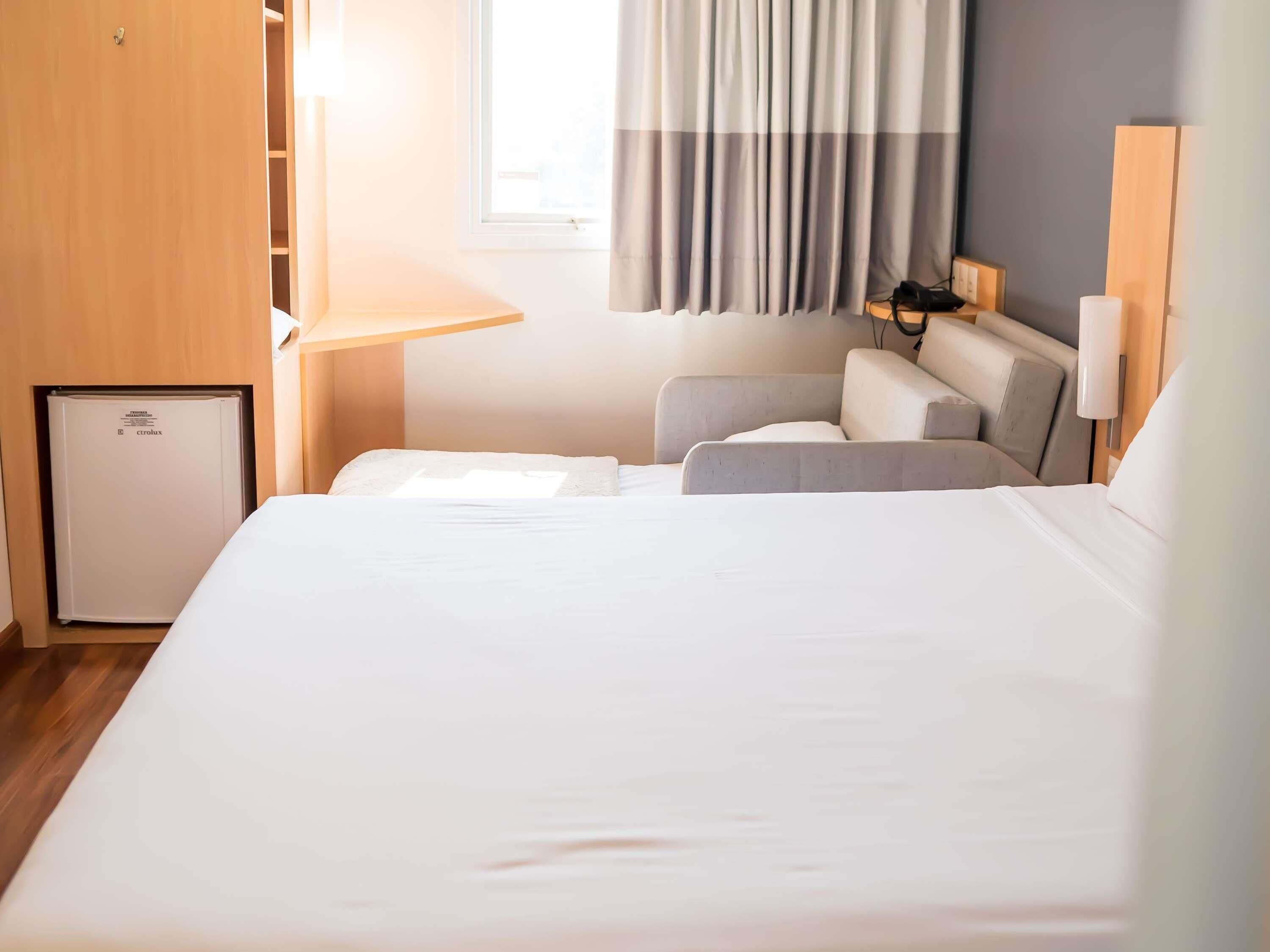 Quarto de hotel com cama, escrivaninha, cadeira, pequeno frigorífico e janela com cortinas.
