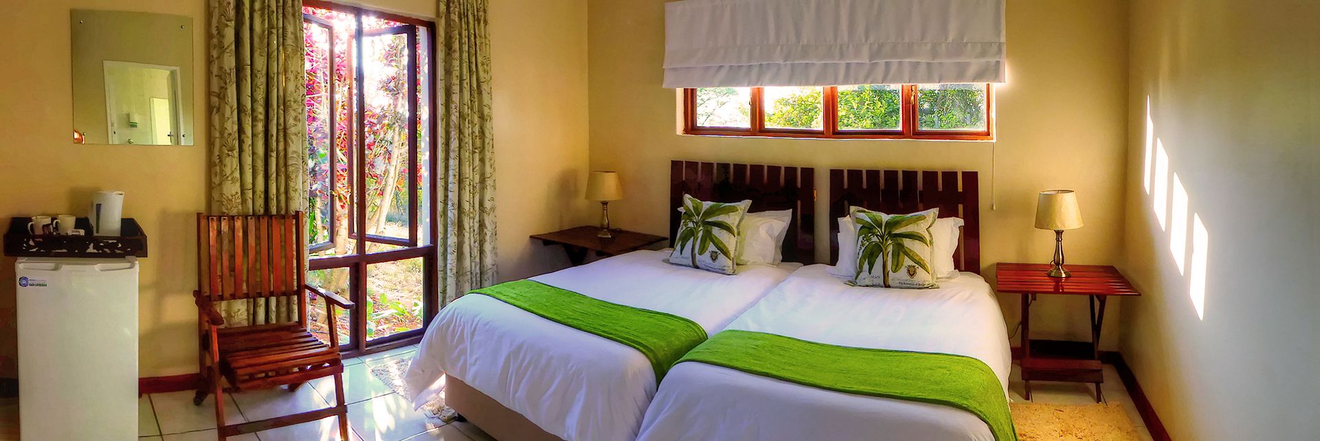 Quarto de hotel com uma cama grande, duas mesas de cabeceira, um criado-mudo, uma cadeira, janela com cortinas e um espelho na parede.