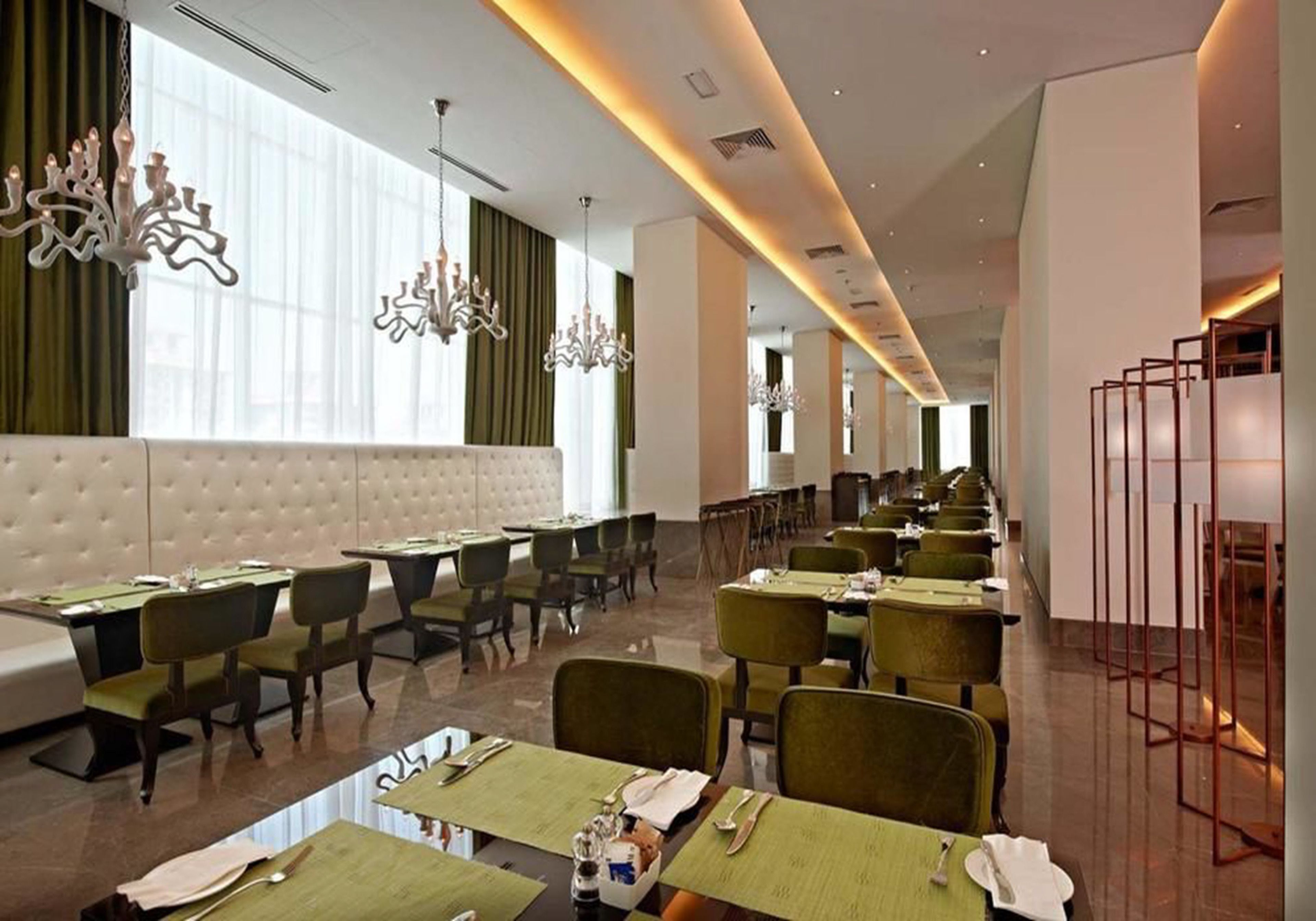 Uma sala de jantar moderna com várias mesas e cadeiras, teto alto com iluminação embutida e grandes janelas que permitem entrada de luz natural.