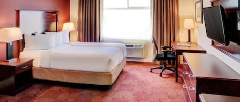 Quarto de hotel com uma cama grande, uma escrivaninha com cadeira, uma televisão e uma janela com cortinas.