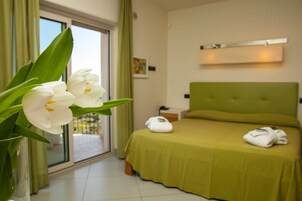 Quarto de hotel com cama, mesa de cabeceira, abajur e vista através de uma porta de vidro.