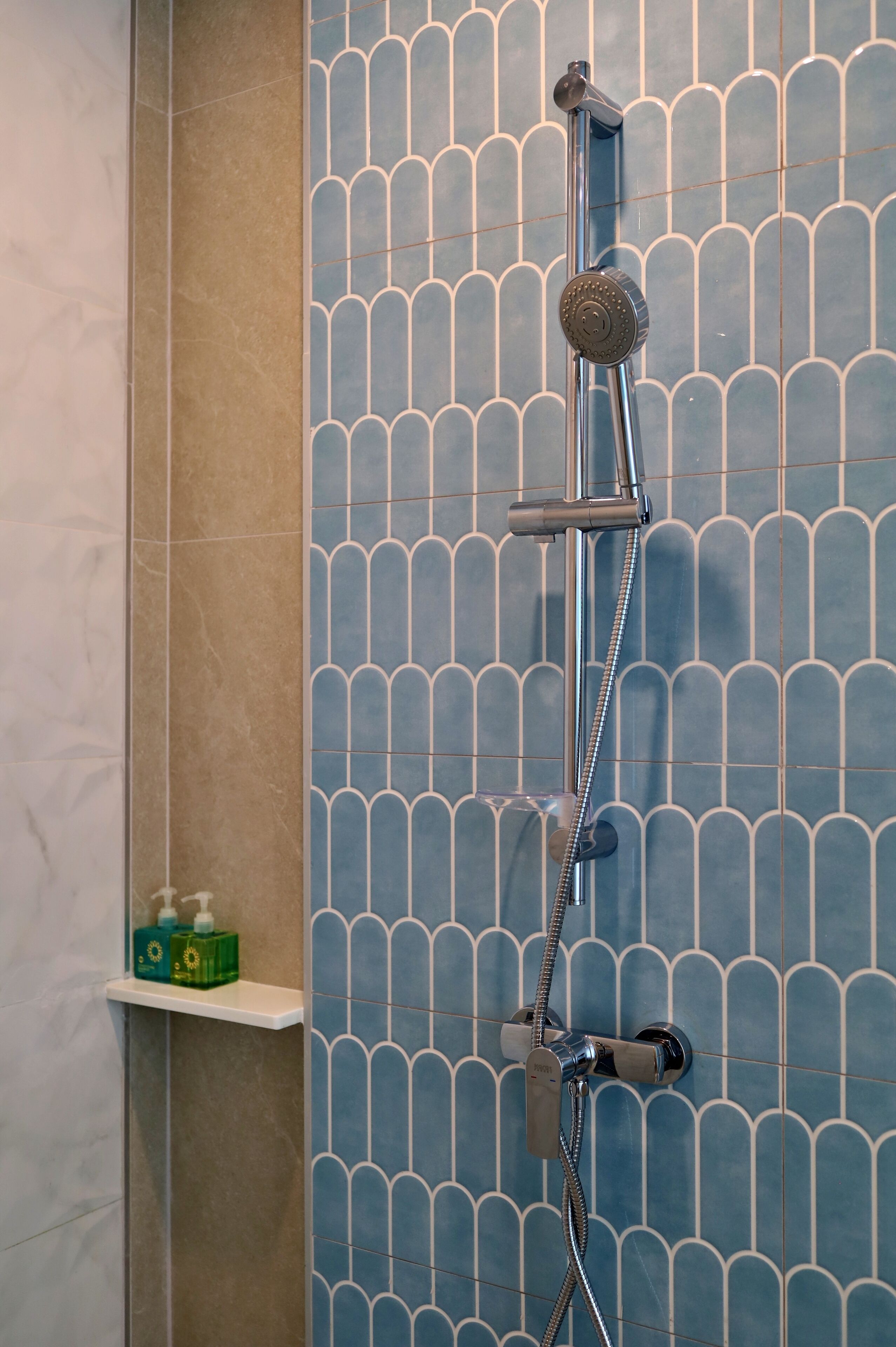 Casa de banho com parede revestida a azulejos azuis, um chuveiro e uma prateleira com artigos de higiene pessoal.