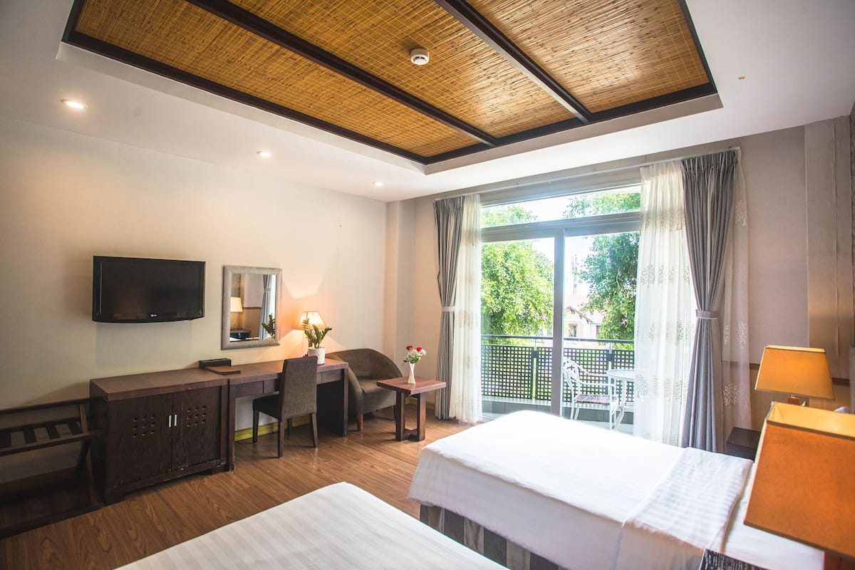 Quarto de hotel com duas camas, uma escrivaninha com cadeira, televisão e varanda com cortinas.