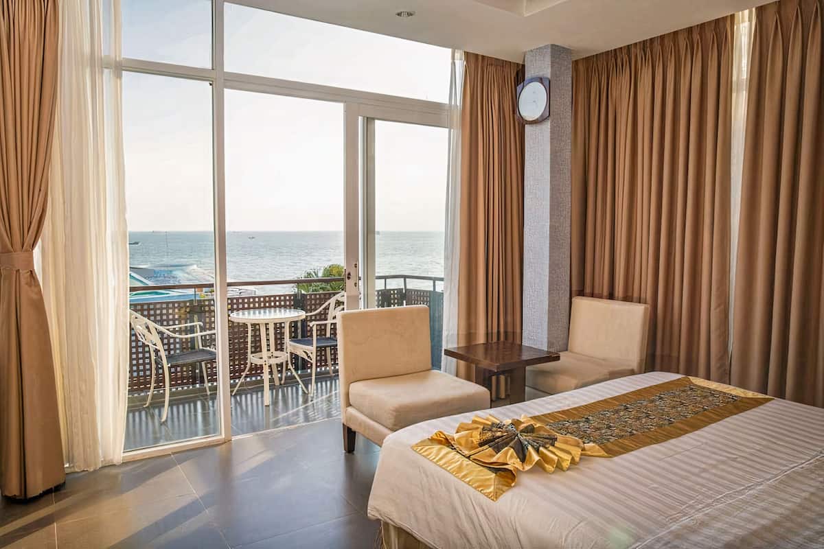 Quarto de hotel com uma grande janela, uma cama com cabeceira decorativa, uma pequena mesa e uma cadeira.