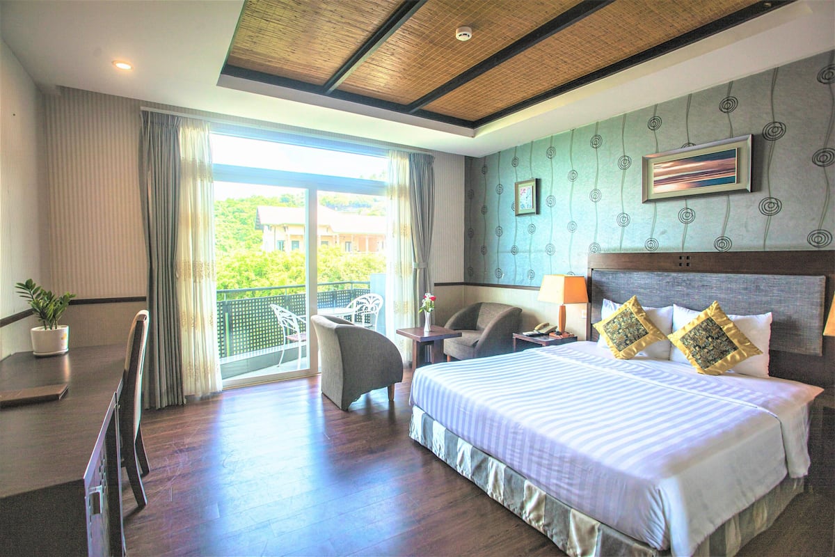 Quarto de hotel com uma cama grande, duas poltronas, uma mesa pequena e varanda com vista para áreas verdes.