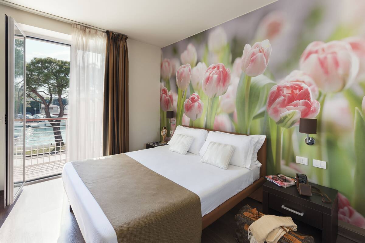 Quarto de hotel com uma cama grande, mesa de cabeceira com abajur, janela com cortinas e um mural de flores na parede.