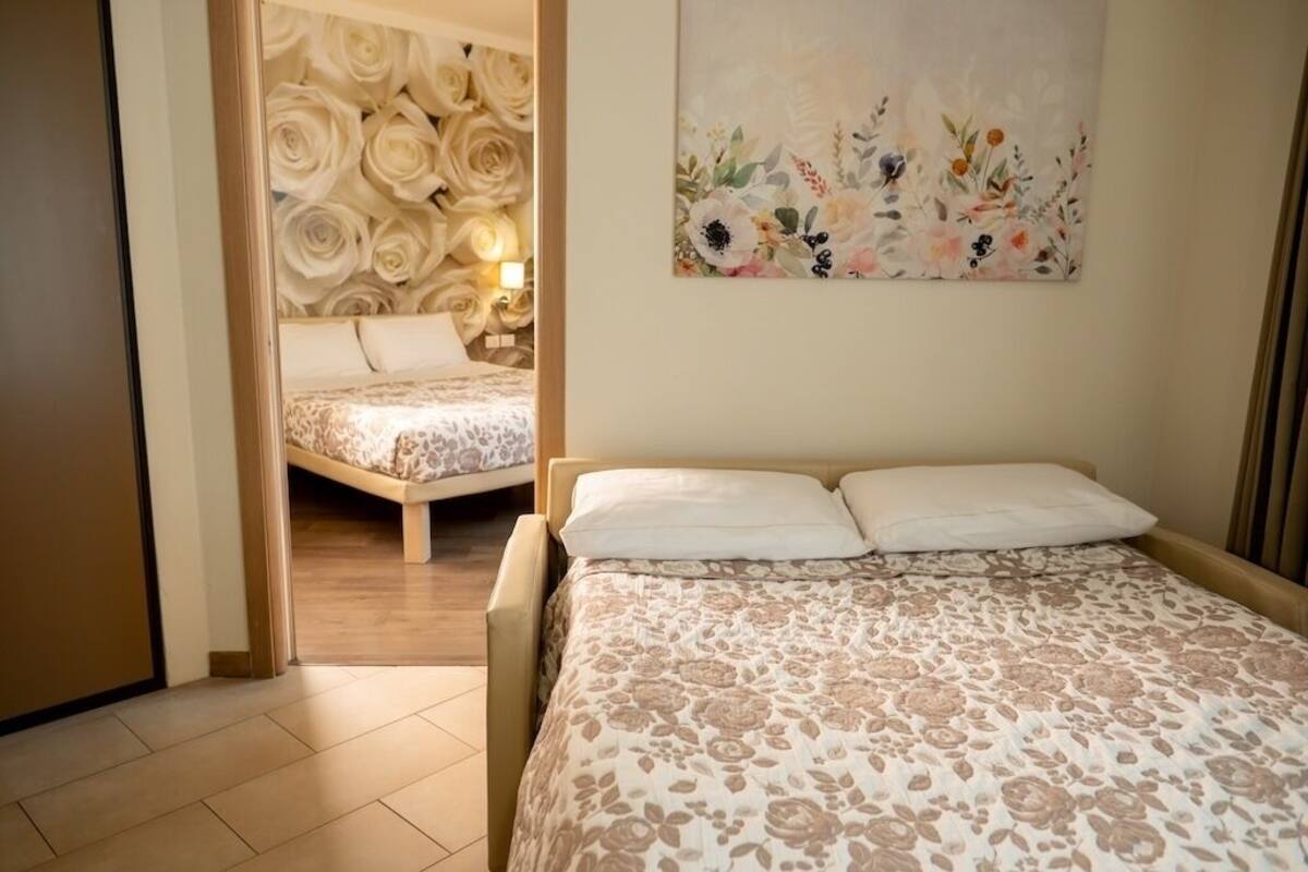 Quarto com cama, mesa de cabeceira, parede decorada com arte floral e porta para outro cômodo.