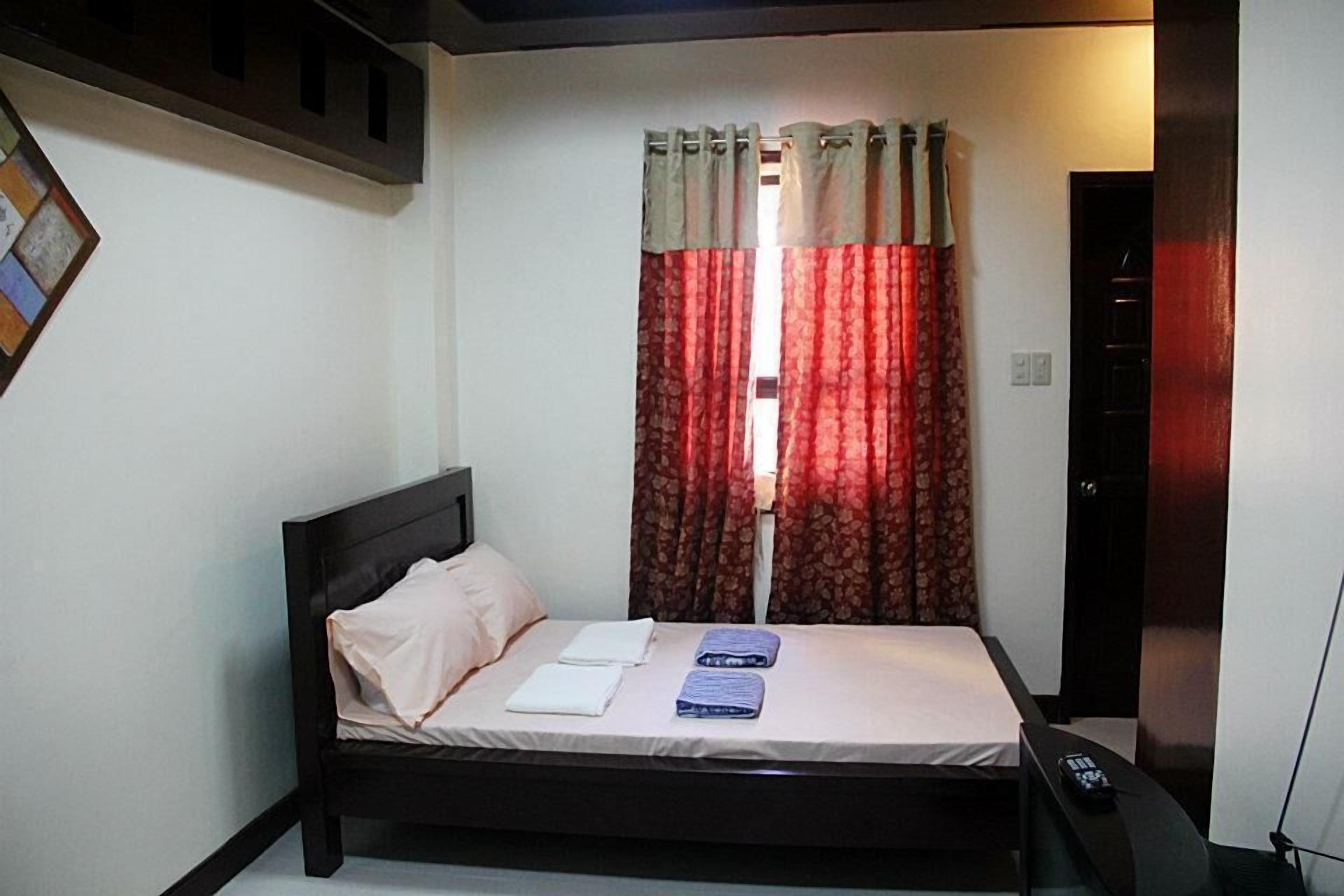Uma cama de solteiro com cabeceira, mesa de cabeceira, controle remoto e janela com cortinas estampadas.