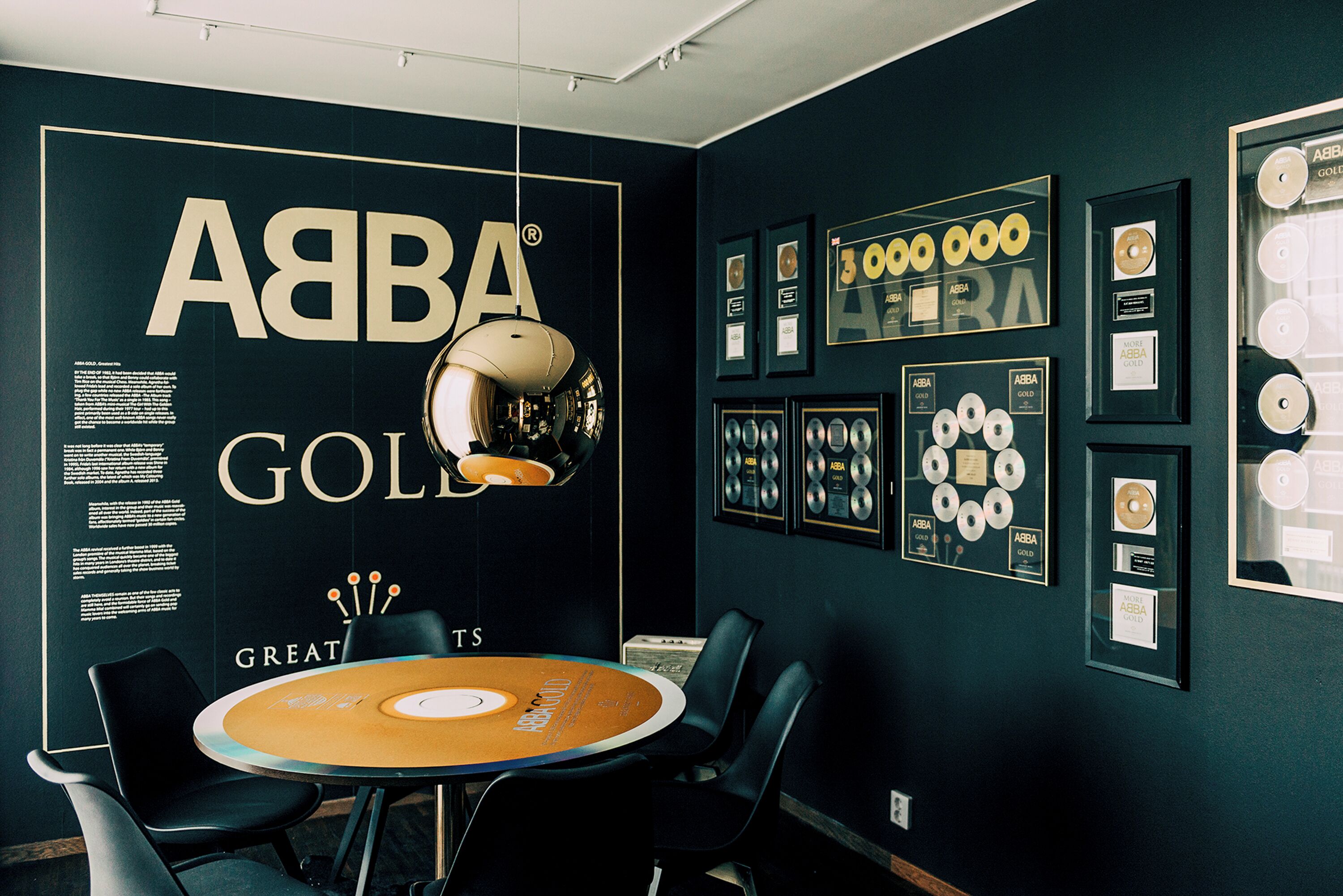 Uma sala de conferências com um grande painel de parede exibindo o logotipo ABBA e prêmios, uma mesa redonda com cadeiras e um lustre.