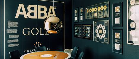 Uma sala de conferências com um grande painel de parede exibindo o logotipo ABBA e prêmios, uma mesa redonda com cadeiras e um lustre.