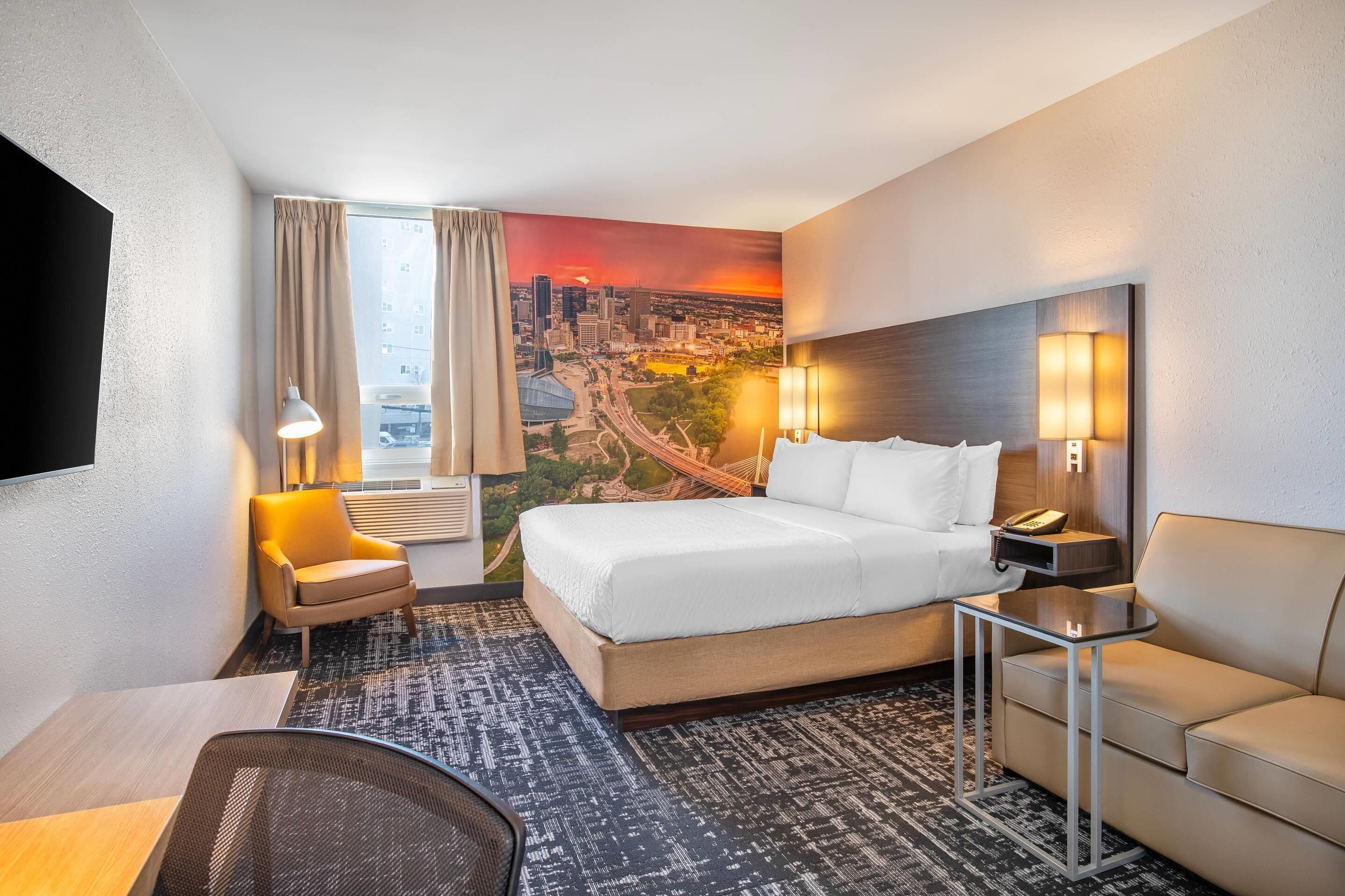 Quarto de hotel com cama, sofá, escrivaninha, cadeira, luminária, mesa de cabeceira e um mural com vista para a cidade na parede.