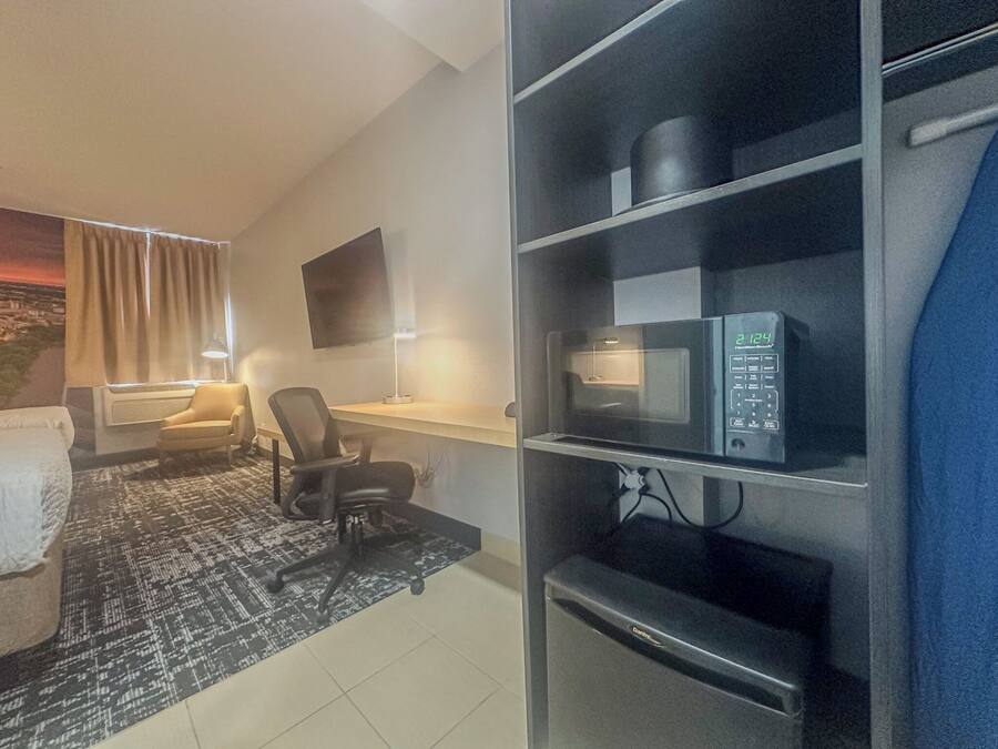 Quarto de hotel com escrivaninha, cadeira, TV de tela plana, micro-ondas e estante de livros.