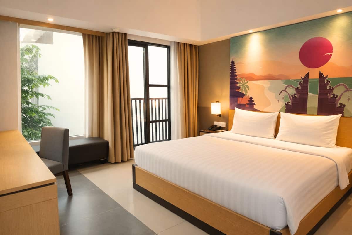 Quarto de hotel com cama, escrivaninha, cadeira, mesa de cabeceira e janela com cortinas.