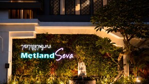 Fachada de prédio com letreiro de neon que diz 'Metland Seva' e uma estátua em frente.