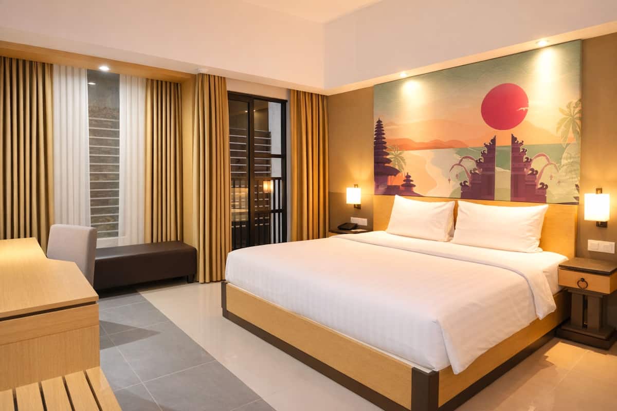 Quarto de hotel com cama, escrivaninha, cadeira, mesa de cabeceira e um quadro na parede.